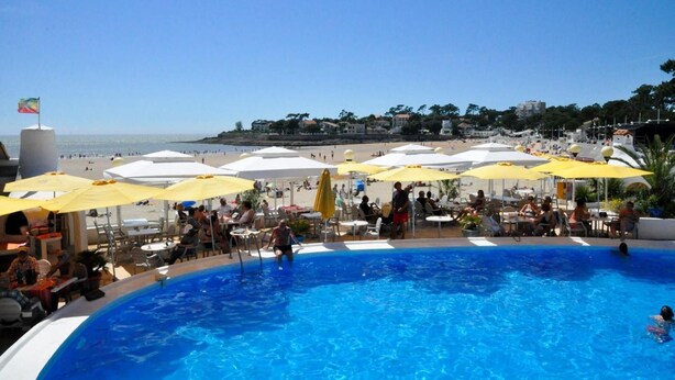 Área da piscina com guarda-sóis, espreguiçadeiras e vista para a praia e o oceano.