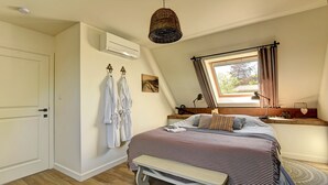 Quarto com cama, um banquinho, janela com cortinas, mesa de cabeceira e ar condicionado fixado na parede.