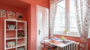 Um quarto com uma estante branca, uma mesa com cadeiras e uma janela com cortinas florais.