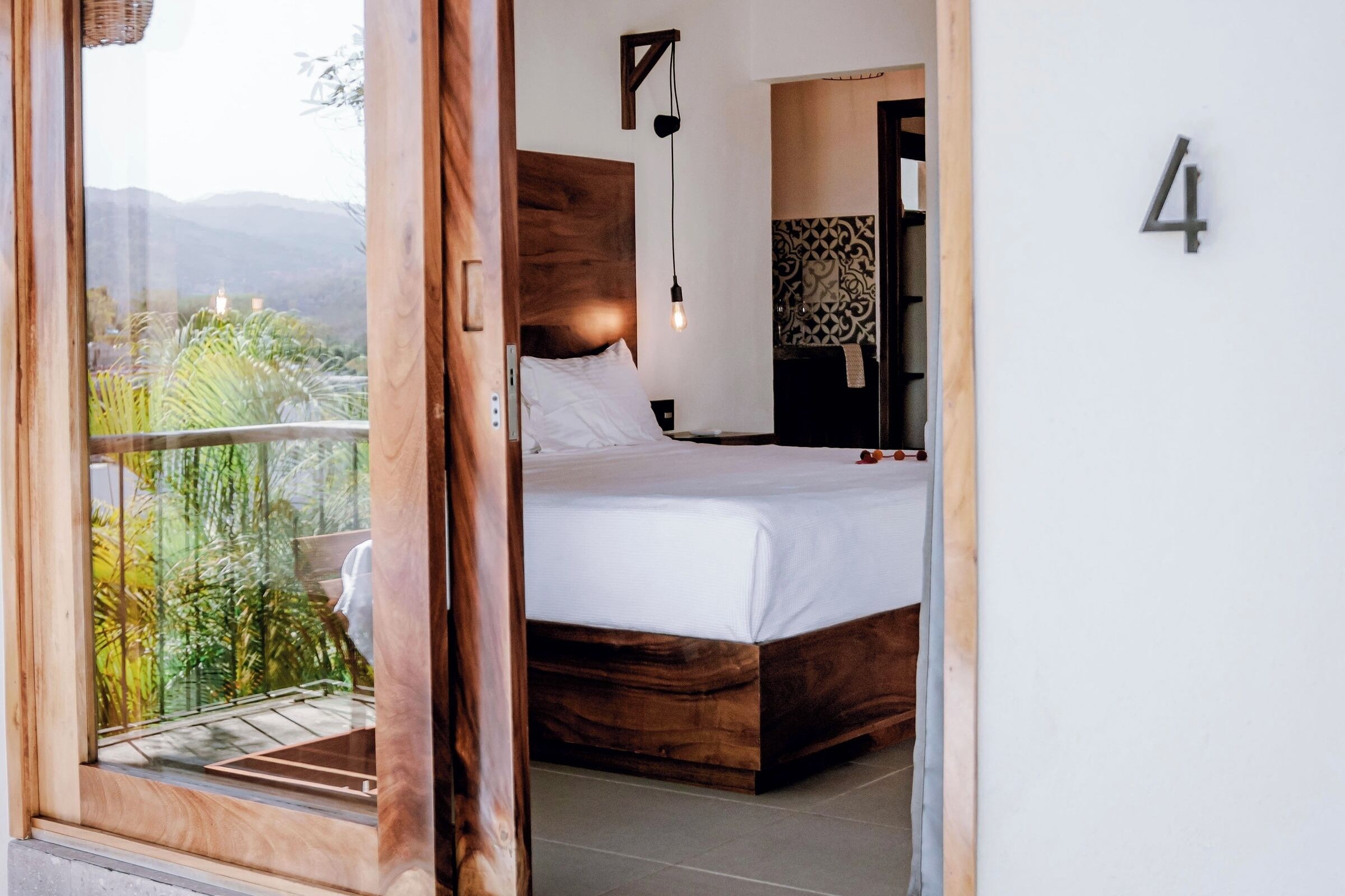 Quarto com cabeceira de madeira, cama com roupa de cama branca, luminária pendente e vista para o verde através de uma porta de vidro.