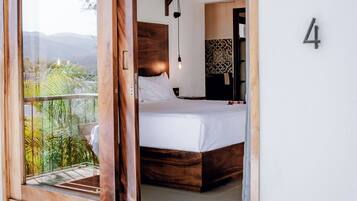 Quarto com cabeceira de madeira, cama com roupa de cama branca, luminária pendente e vista para o verde através de uma porta de vidro.