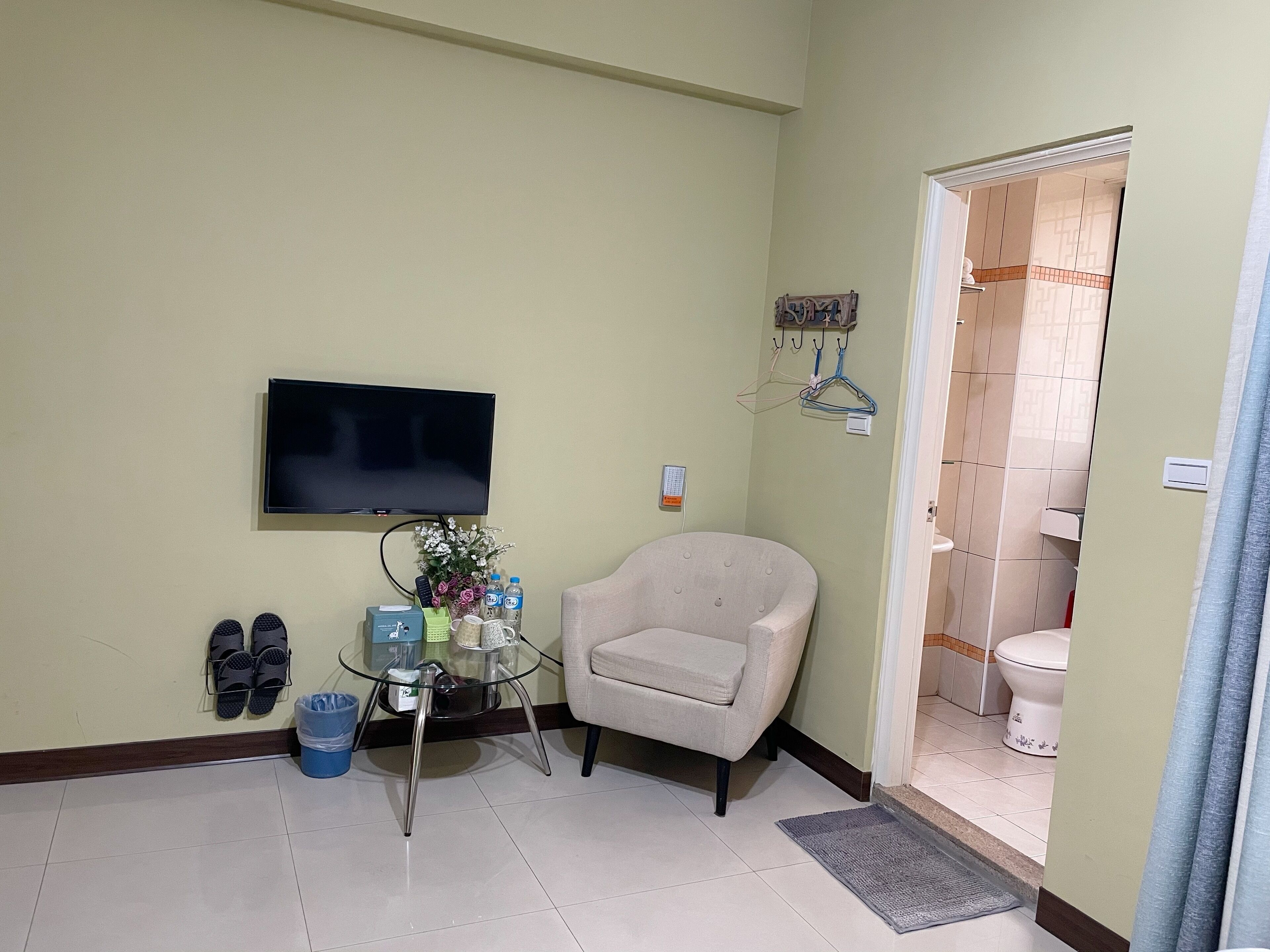 Sala de estar com televisão, uma cadeira, uma mesinha com objetos e a porta de um banheiro.