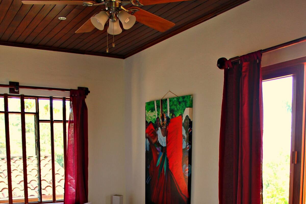 Quarto de hotel com cama, ventoinha no teto, um quadro e janela com cortinas.