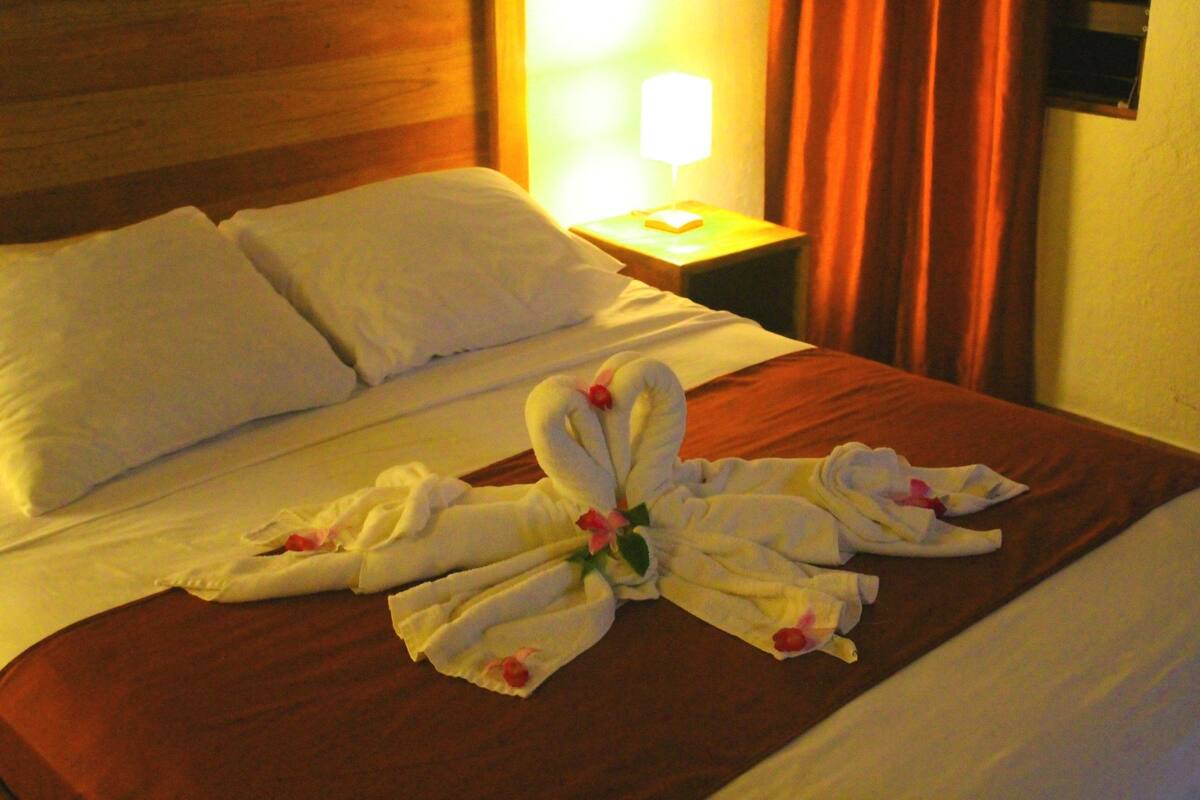 Cama bem arrumada com lençóis brancos e uma toalha dobrada em forma de laço decorativo, com uma flor.