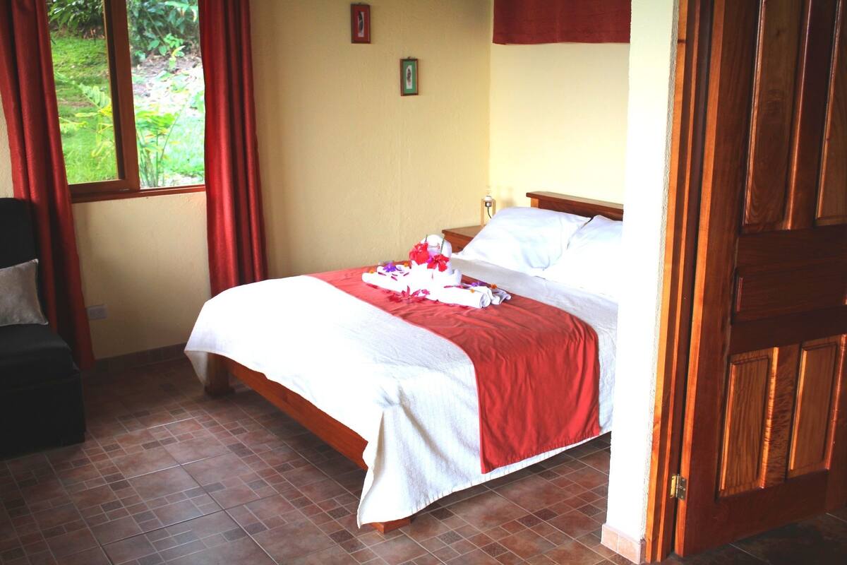 Cama bem arrumada com colcha branca e vermelha, cabeceira de madeira e uma pequena mesa de cabeceira com abajur.