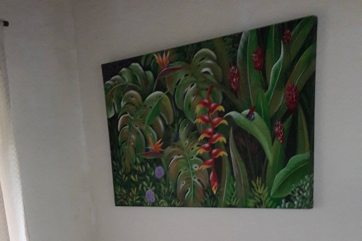 Quarto de hotel com cama de madeira, roupa de cama branca, colcha verde, pintura de flores tropicais na parede e duas luminárias de cabeceira.