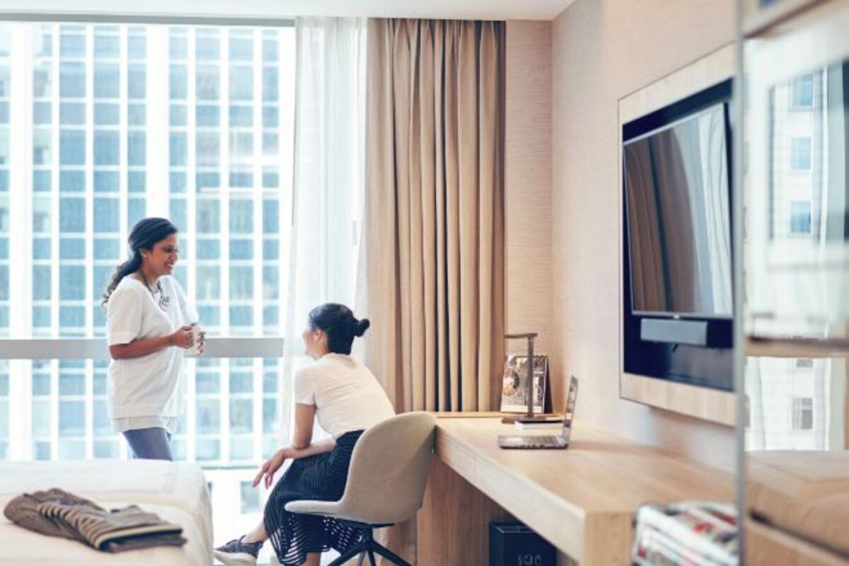 Quarto de hotel com uma janela grande, uma mesa com televisão de tela plana e uma cama com cobertor.