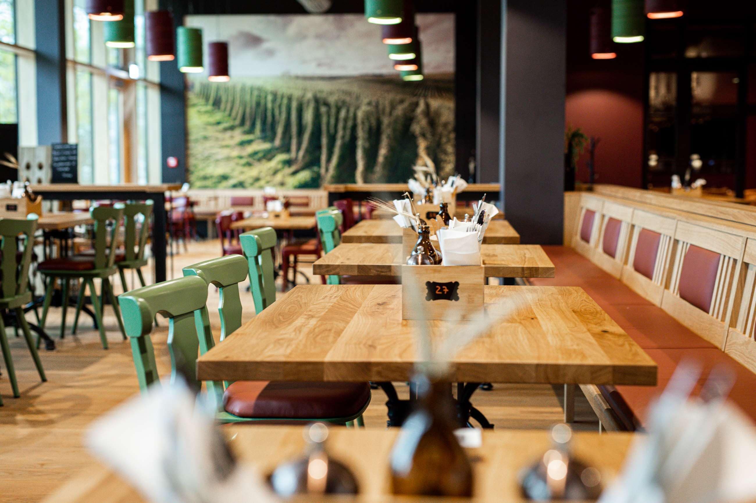 Um restaurante com mesas de madeira e cadeiras verdes, um mural na parede e iluminação pendente.