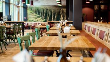 Um restaurante com mesas de madeira e cadeiras verdes, um mural na parede e iluminação pendente.