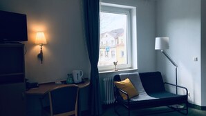 Quarto de hotel com escrivaninha, cadeira, sofá, duas luminárias, radiador e janela com vista para edifícios.