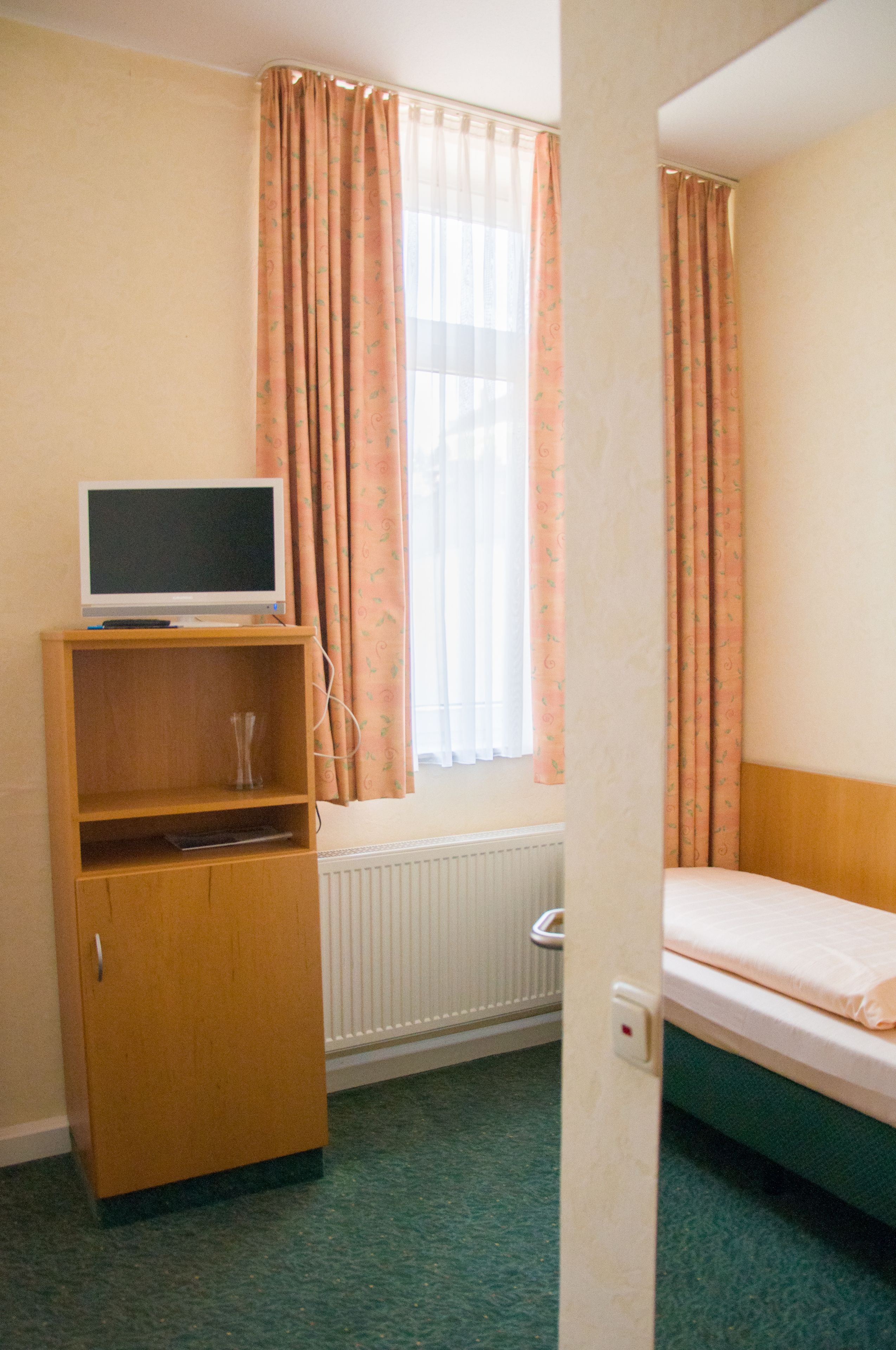 Quarto de hotel pequeno e individual, com uma cama, um armário de madeira com televisão, uma janela com cortinas e um radiador.