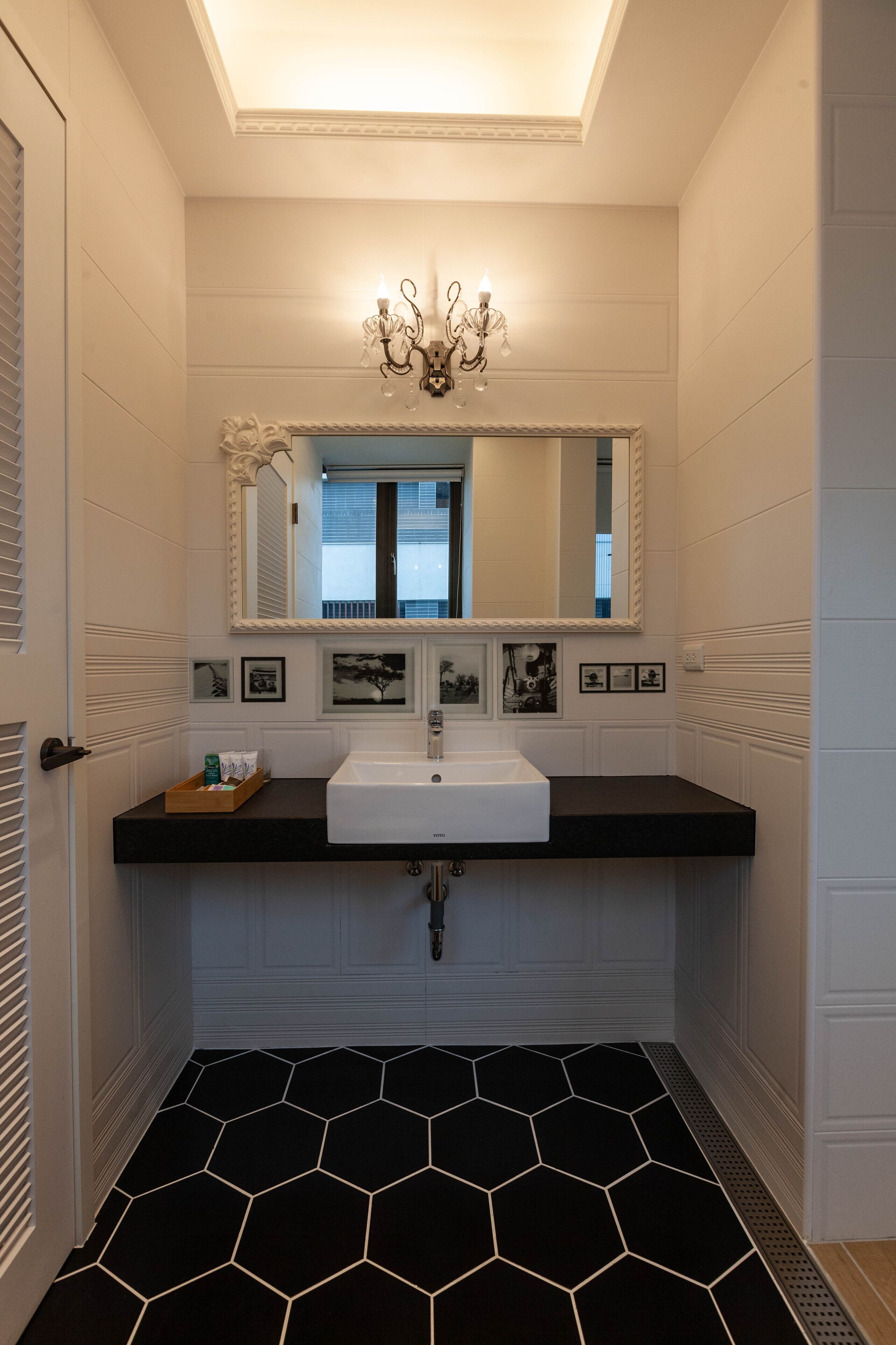 Um banheiro com toucador branco, ladrilhos hexagonais pretos no chão, um candelabro e quadros nas paredes.