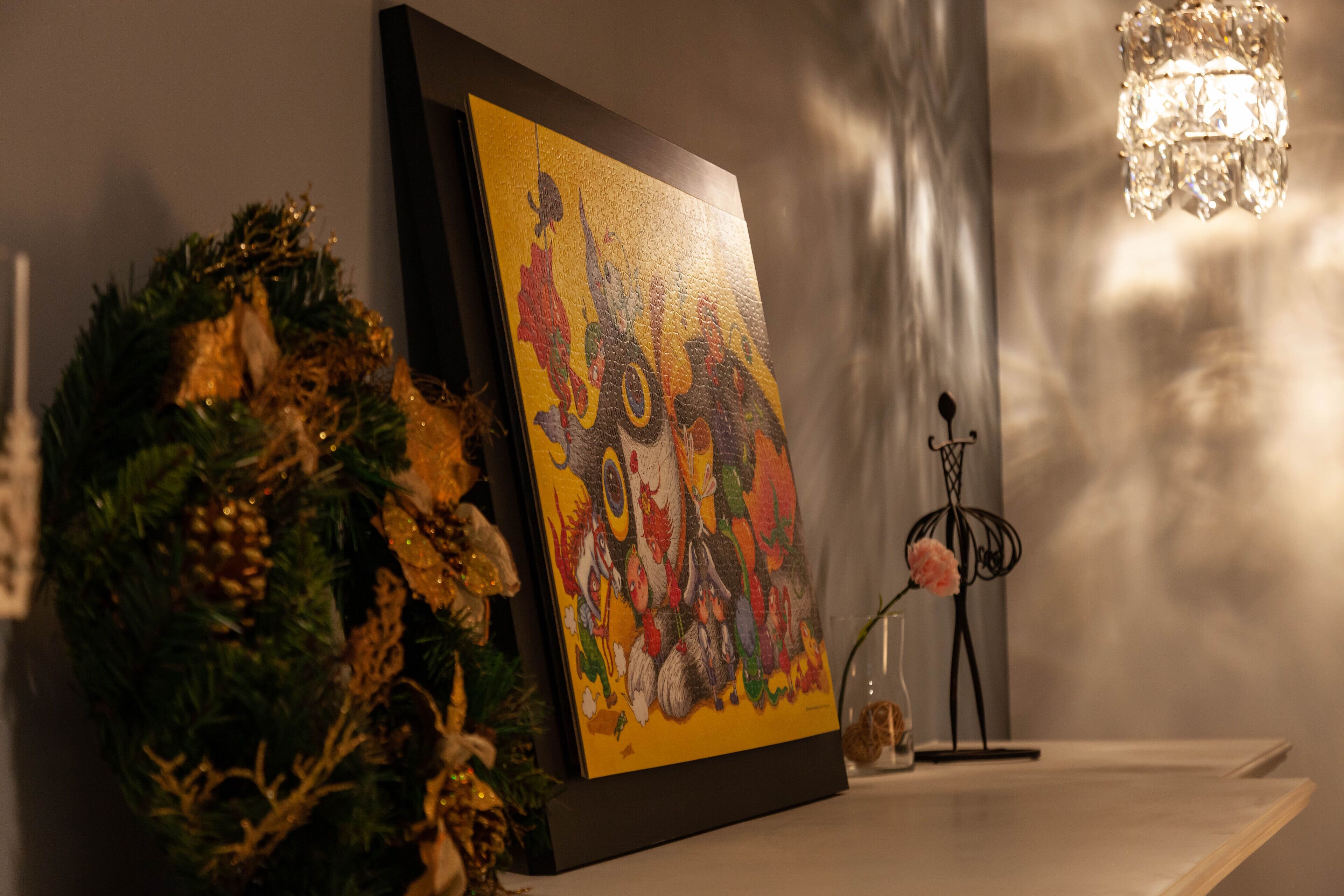 Uma obra de arte emoldurada sobre uma superfície branca, uma coroa decorativa e um castiçal com uma flor rosa.