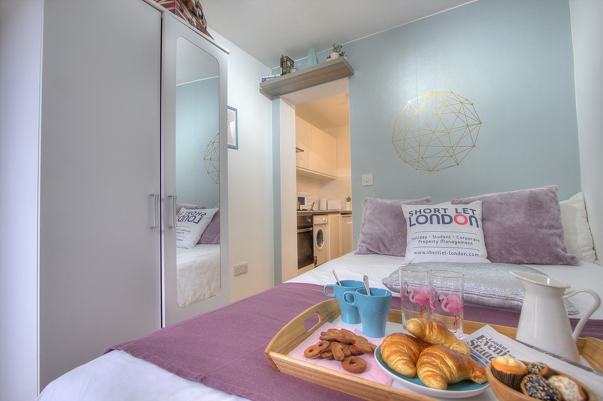 Quarto com cama, bandeja com croissants e um muffin, cafeteira e travesseiro com a inscrição 'SHORT LET LONDON'.