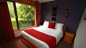 Quarto com uma cama grande, mesas de cabeceira de madeira, colcha vermelha e branca, vista para o verde através de uma janela e um tapete estampado.