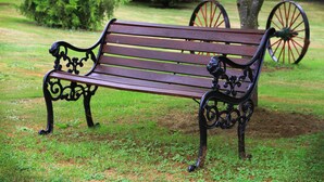 Um banco de madeira com pernas de metal ornamentadas numa área gramada, com árvores e uma grande roda ao fundo.