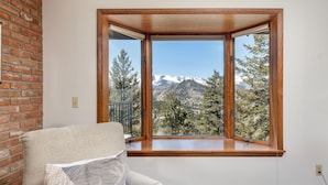 Quarto com grande janela que oferece vista para as montanhas, parede de tijolos e piso de madeira.