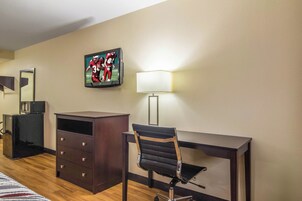 Quarto de hotel com escrivaninha, cadeira, cômoda e televisão de tela plana fixada na parede.