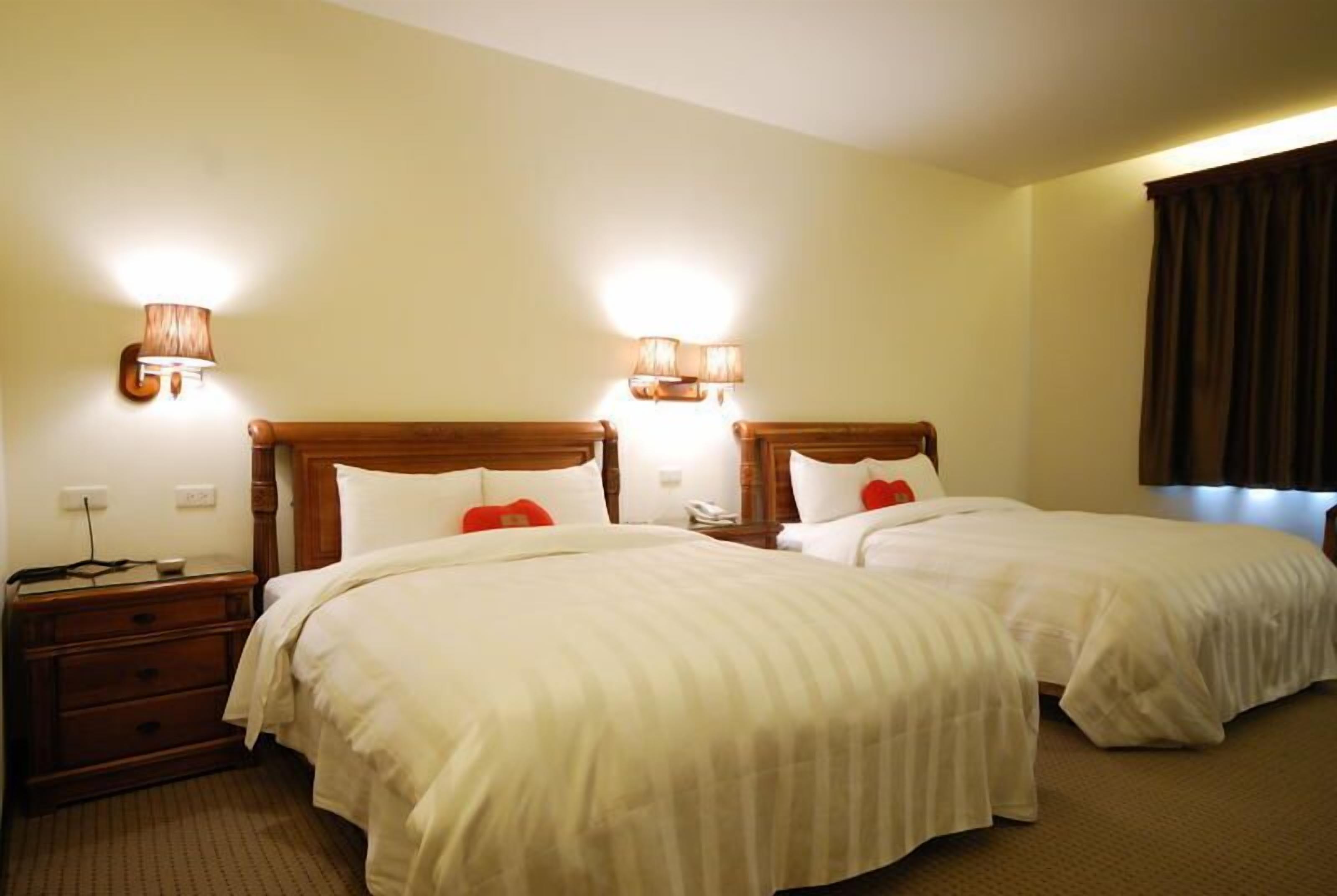 Quarto de hotel com duas camas, cada uma com roupa de cama branca e almofadas vermelhas. Há luminárias fixadas na parede em ambos os lados das camas.