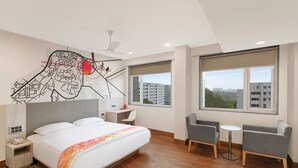 Quarto de hotel moderno com uma cama grande, uma escrivaninha, duas cadeiras, uma mesinha e uma janela ampla com vista para a cidade.