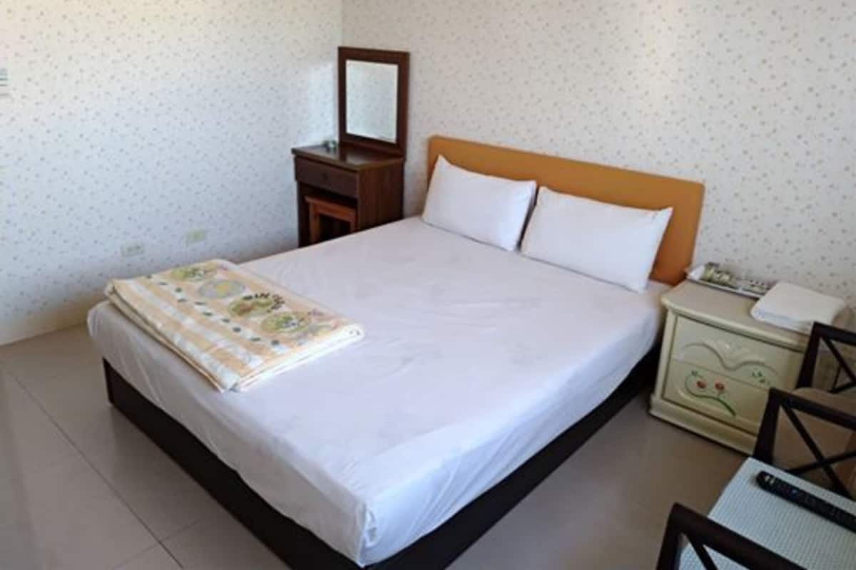 Cama bem arrumada com lençóis brancos e um manta decorativa, mesa de cabeceira de madeira com espelho, e pequena mesa lateral com abajur.