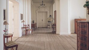 Um corredor com piso de madeira, porta branca e um candelabro. Cadeiras e uma mesa com abajur estão do lado esquerdo.