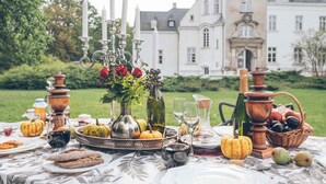 Mesa posta para refeição com vários pratos, um vaso de flores e uma garrafa de vinho, com um edifício grande ao fundo.