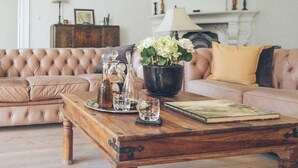 Uma sala de estar com um sofá de couro, uma mesa de centro de madeira com um livro e copos, um vaso com flores e uma lareira ao fundo.
