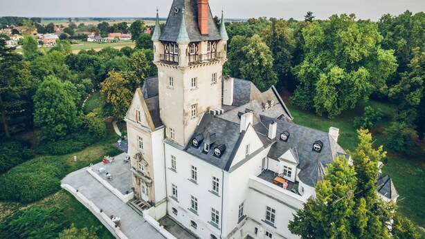Um edifício grande e histórico com uma torre imponente e várias alas, rodeado por vegetação exuberante e uma aldeia ao longe.