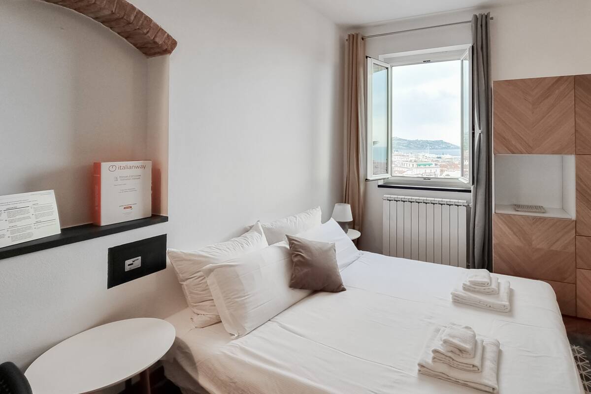 Quarto de hotel com cama, mesa de cabeceira, luminária, guarda-roupa e janela com vista para a cidade.