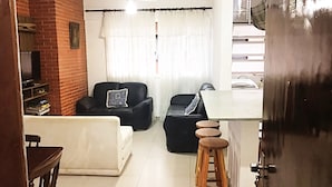 Uma sala de estar com lareira de tijolos, um sofá de couro preto, um sofá branco e uma mesa de jantar de madeira com banquetas.
