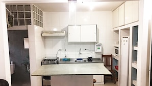 Cozinha compacta com ilha central, fogão a gás, pia e armários brancos. Três banquetas de madeira estão em frente à ilha.