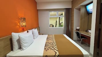 Quarto de hotel com cama, escrivaninha, televisão e janela com vista para o verde.