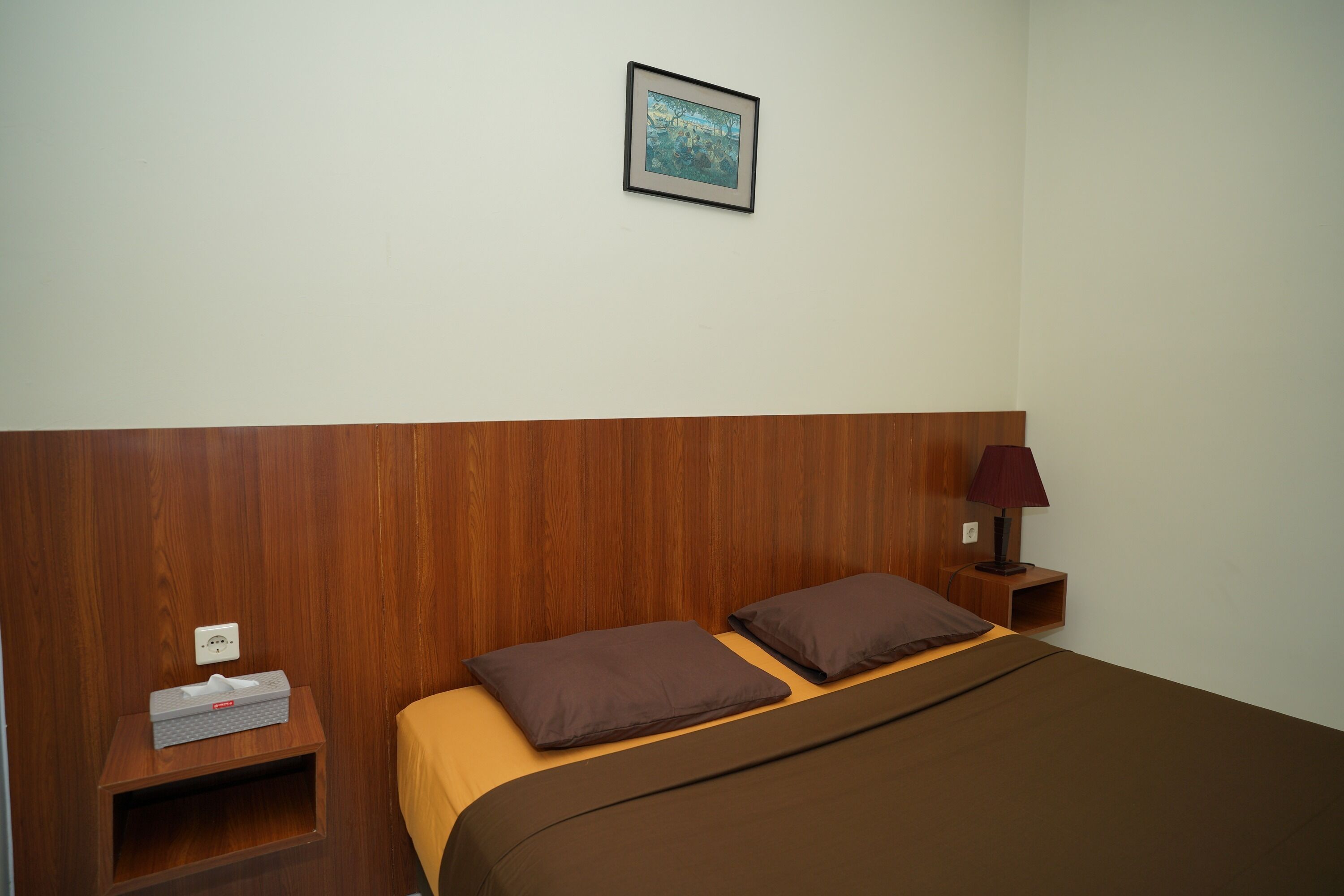 Quarto de hotel com cabeceira de madeira, cama com roupa de cama marrom, mesa de cabeceira com abajur e quadro na parede.