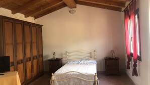 Quarto com teto de madeira, cama com cabeceira branca, duas mesas de cabeceira, um guarda-roupa e uma janela com cortinas vermelhas.