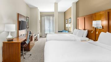 Quarto de hotel com duas camas, uma mesa de trabalho com cadeira, televisão e banheiro visível por uma porta.