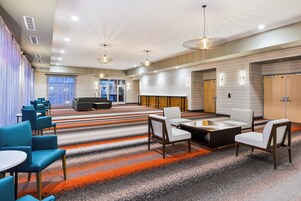 Um saguão de hotel com design moderno, apresentando uma área de estar com mesas e cadeiras, um balcão de recepção e uma grande janela.