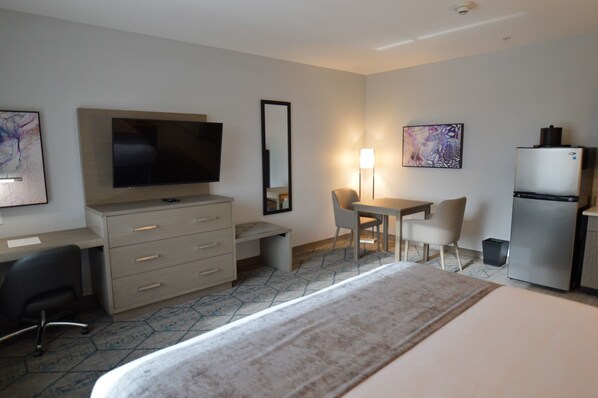 Quarto de hotel com cama, escrivaninha, cadeira, televisão, frigorífico e obras de arte nas paredes.