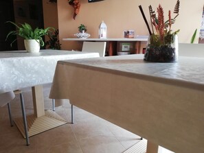 Uma área de refeições com mesas cobertas por toalhas brancas, uma planta em vaso e um vaso decorativo com flores vermelhas.