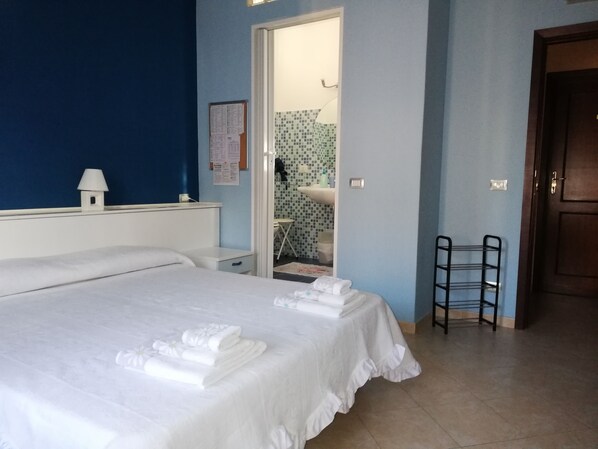 Quarto de hotel com duas camas, uma mesa pequena, uma cadeira, uma porta e um banheiro visível através de uma porta aberta.