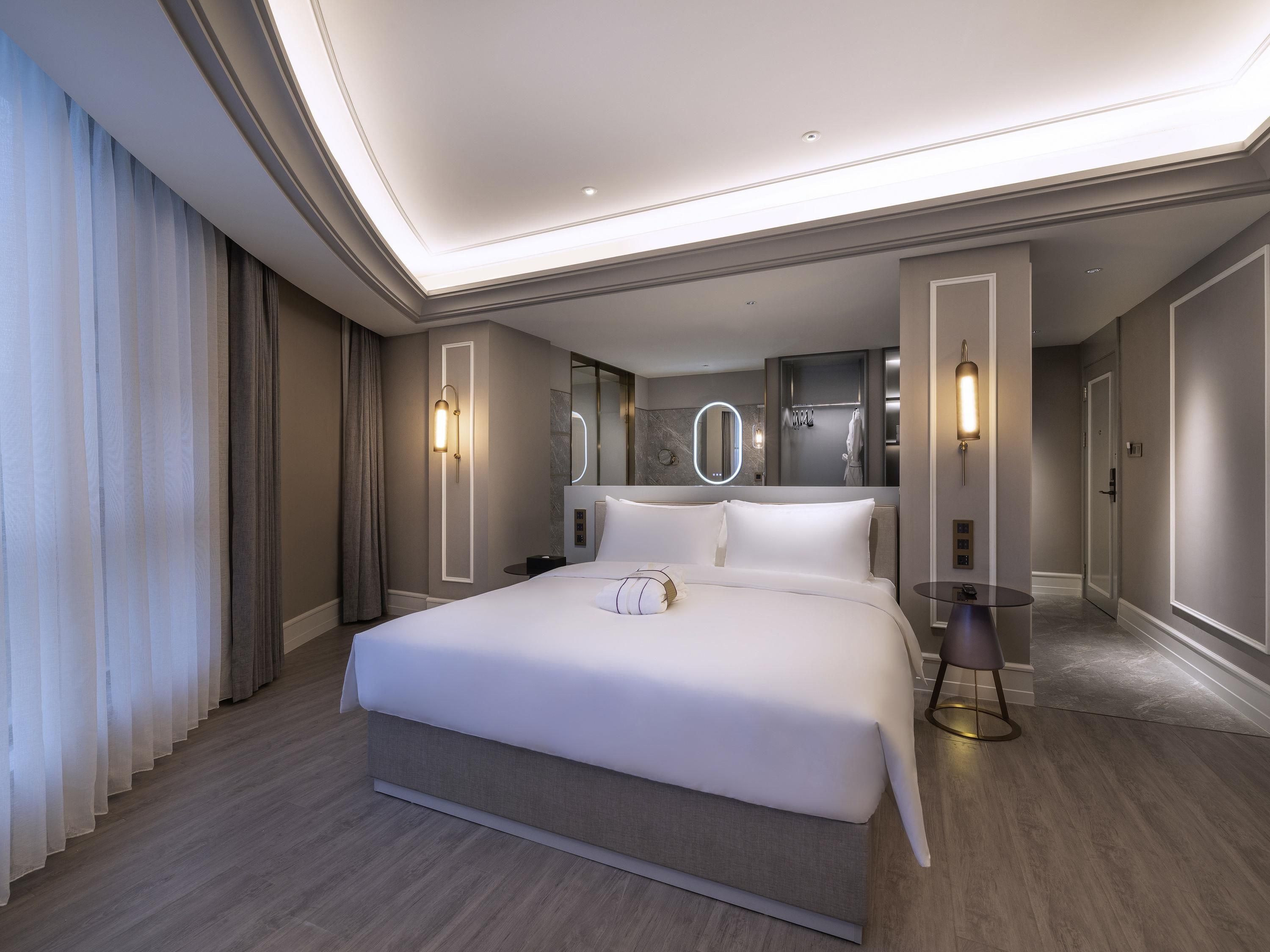 Quarto de hotel moderno com uma cama grande, uma mesa de cabeceira, um espelho e um banheiro visível por uma porta aberta.