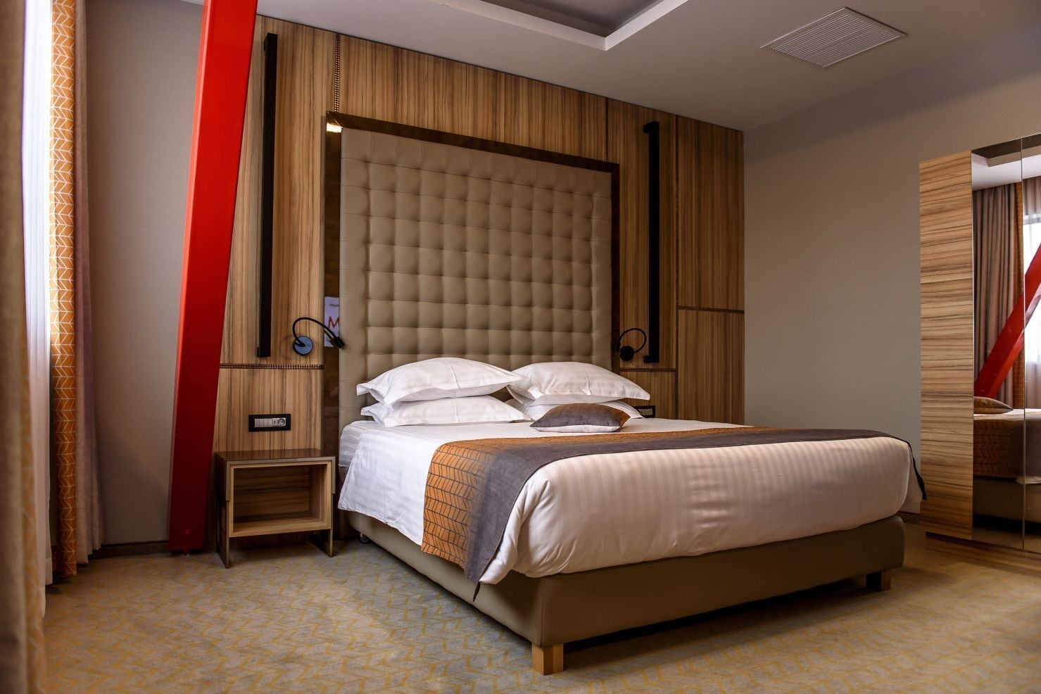 Quarto de hotel com uma cama grande, cabeceira de madeira, mesa de cabeceira e espelho.