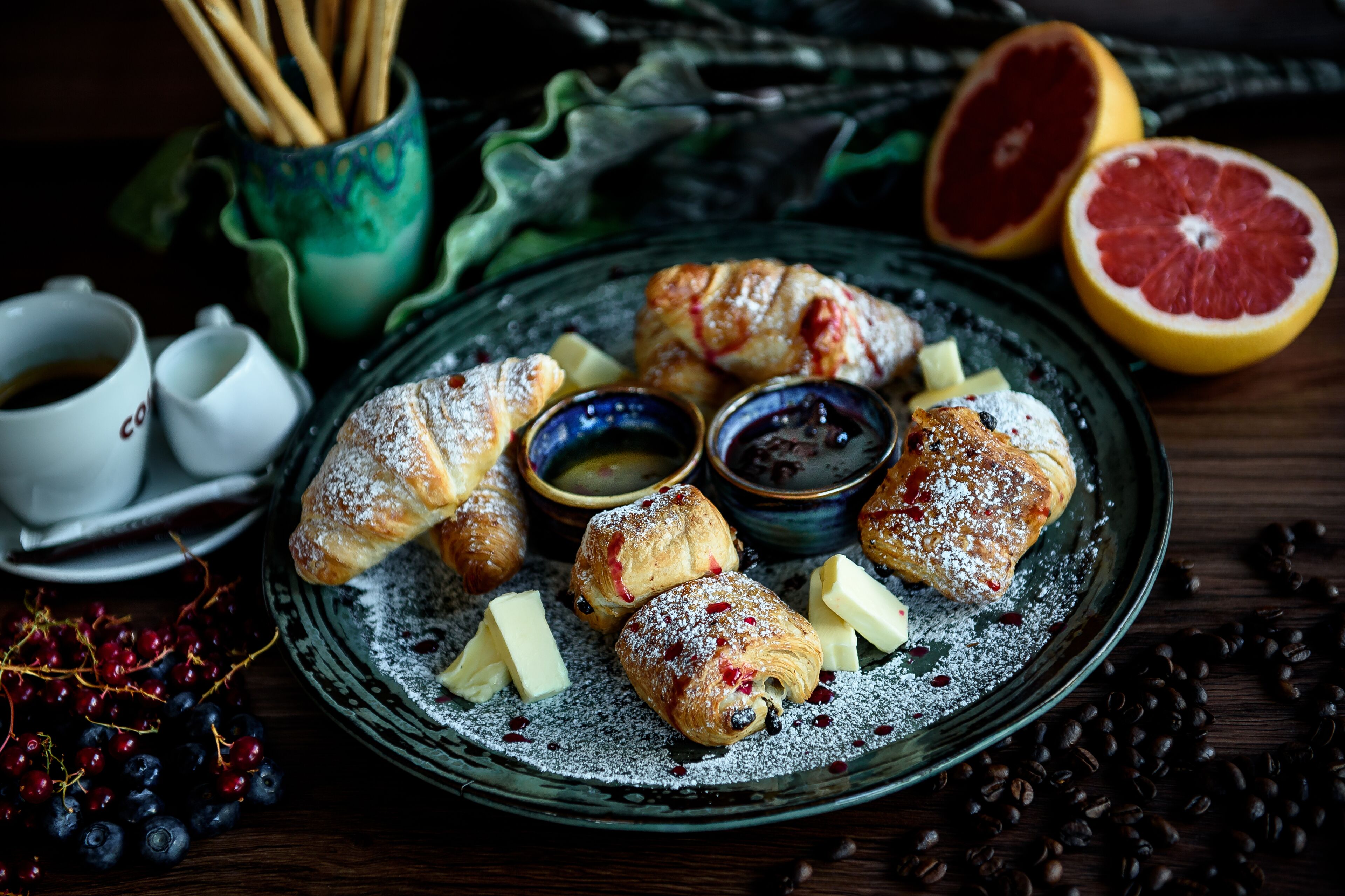 Um prato de doces com açúcar em pó, manteiga e frutas vermelhas, acompanhado de xícaras de café e meia toranja.