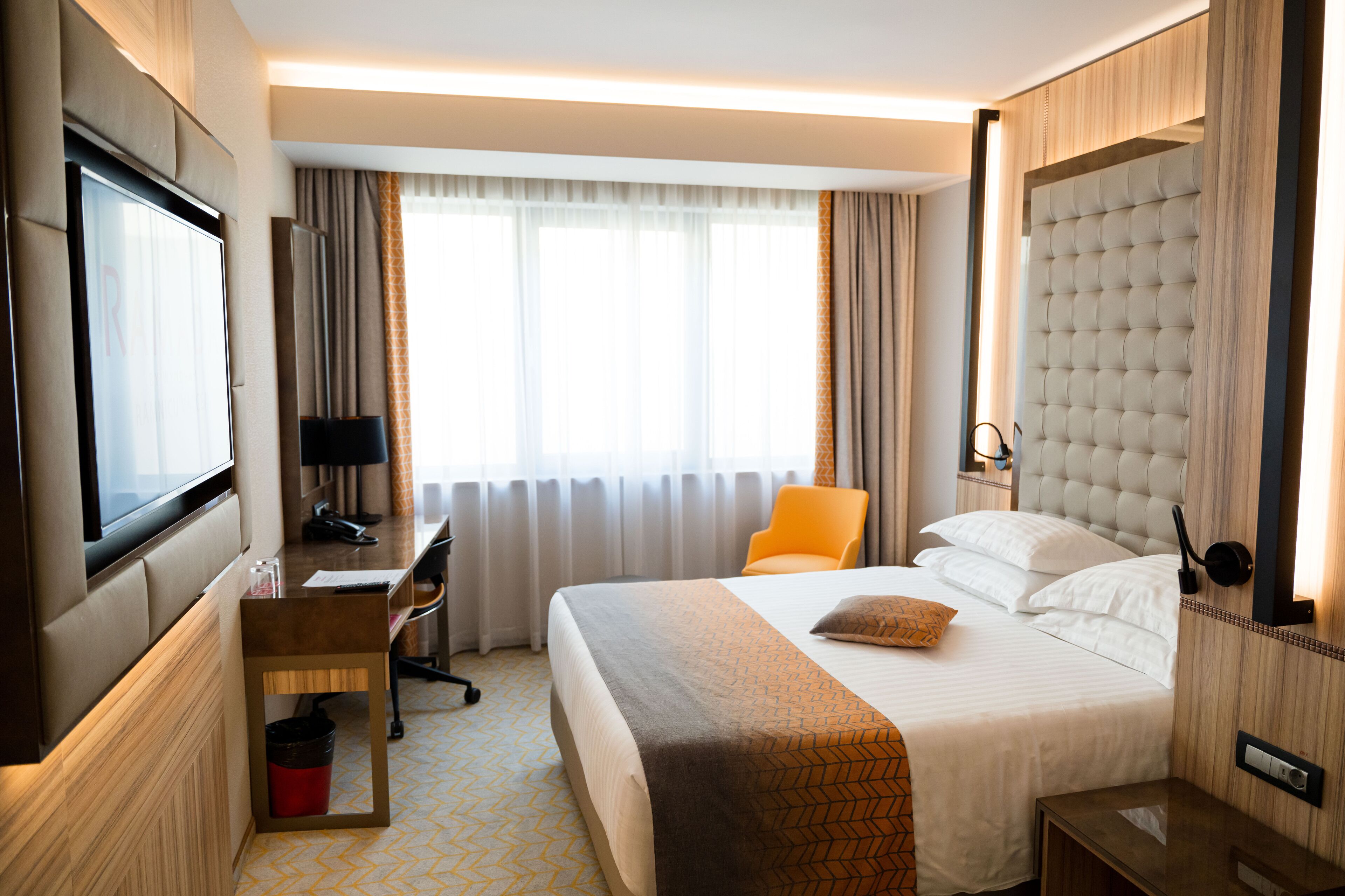 Quarto de hotel com cama, escrivaninha, cadeira, televisão e janela com cortinas.