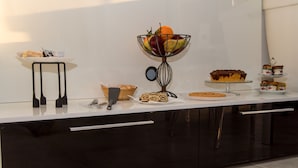 Uma bancada de cozinha com uma fruteira, pão, um prato de biscoitos e um bolo em um suporte.