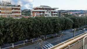 Um edifício residencial de vários andares com varandas, uma rua com faixa de pedestres e uma fileira densa de árvores ao primeiro plano.