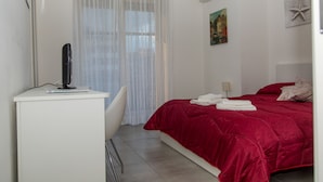 Quarto com cama, escrivaninha com computador, cadeira, janela com cortinas leves e ar condicionado.