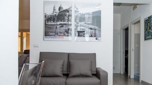 Uma sala de estar com um sofá cinzento, dois cartazes na parede e um corredor que dá para outros cômodos.