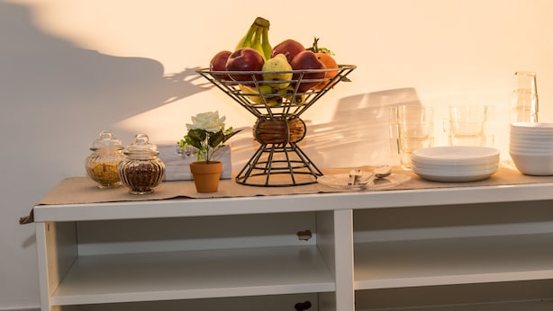 Uma mesa branca com uma cesta de frutas, potes de vidro, uma planta em vaso e pratos empilhados.