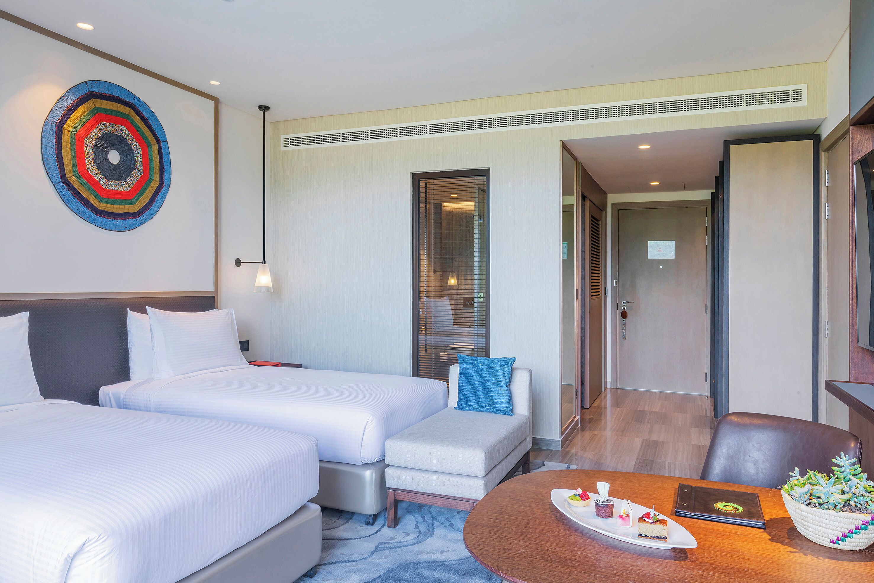 Quarto de hotel com duas camas, um espaço de estar pequeno, uma mesa de jantar com um laptop e uma tigela decorativa, além de uma obra de arte colorida na parede.